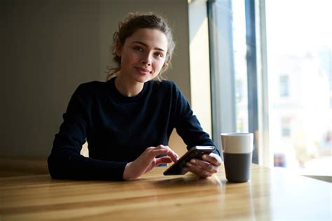 맛 있는 커피 실내 나무 테이블에 앉아 현대 스마트폰 새로운 모바일 응용 프로그램을 설치 하는 동안 카메라를 보고 검은 셔츠 입은 매력적인 여성 학생의 초상화 Stem
