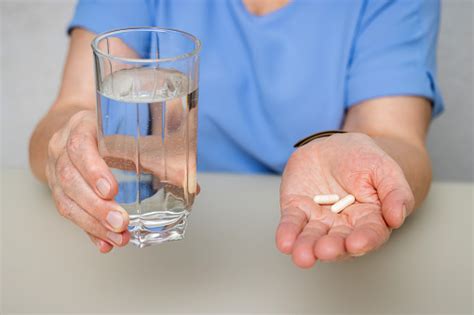 테이블에 앉아 오래된 주름진 손에 흰색 캡슐 비타민 또는 알약을 들고 치료와 물 유리 건강 관리 및 의학 개념을 들고 있는 고위 여성 60 69세에 대한 스톡 사진 및 기타