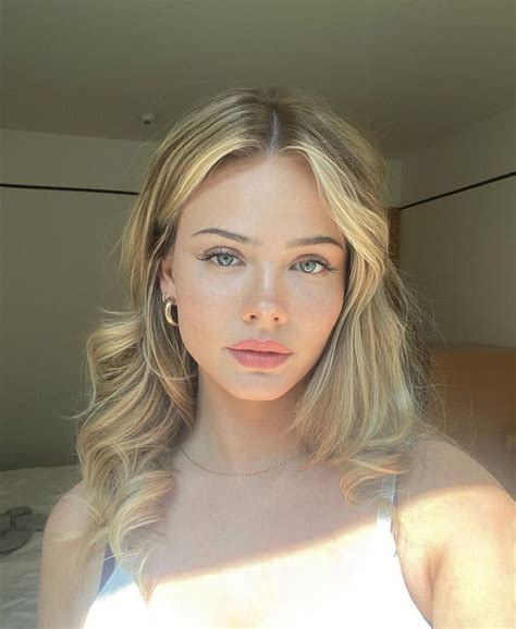 A Woman With Blonde Hair And Blue Eyes Posing For A Photo In Front Of A Bed