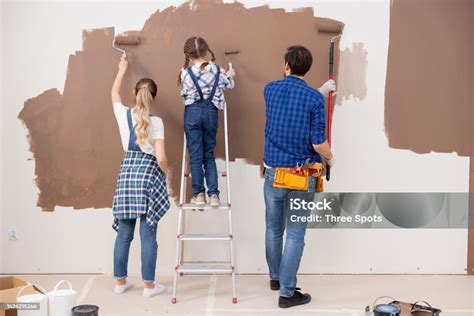 뒤에서 세 사람이 갈색으로 벽을 그렸다 어린 소녀가 사다리 위에 서서 부모님을 도와 수리를 기꺼이 도와줍니다 모두가 함께 시간을 보내는 것을 즐긴다 집 개조에 대한 스톡