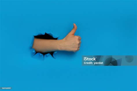 여성 손 제스처 확인 승인 기호는 엄지 손가락을 유지 파란색 종이 배경을 통해 무언가를 돌파하도록 권장합니다 공간을 복사합니다 벽에 구멍 손이 엄지 손가락을 올리는 제스처는