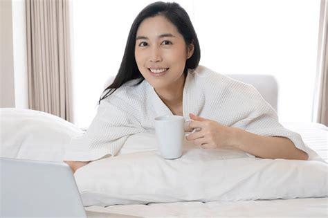 침대에 노트북에서 작업 목욕 가운에 행복 아름다운 아시아 여성 여자는 침실에서 커피 한 잔과 함께 침대에 누워 침대에 어린 소녀는 노트북 컴퓨터를 사용하고 커피를 마십니다
