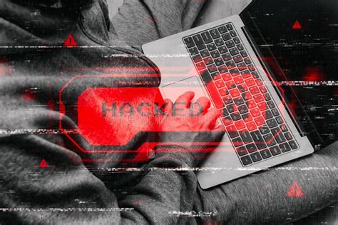 Hacker In A Hoodie Holds A Laptop With A Red Hacked Warning And Binary Code Overlay Symbolizing