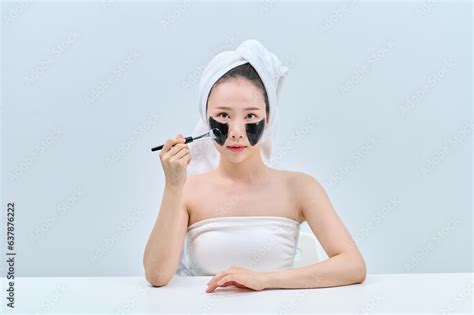 아시아 한국의 젊은 여자 모델이 앉아서 머리에 흰색 수건을 두르고 쇄골과 어깨라인이 보이는 브라탑을 입고 검정색 실리콘 붓을 들고 얼굴에 검은색 모델링 팩을 바르고 있다