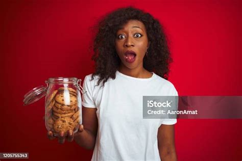 고립 된 빨간 배경 위에 쿠키 항아리를 들고있는 젊은 아프리카 계 미국인 여성은 깜짝 얼굴로 충격에 무서워하고 두려움과 두려움표현으로 흥분했습니다 감정에 대한 스톡 사진 및