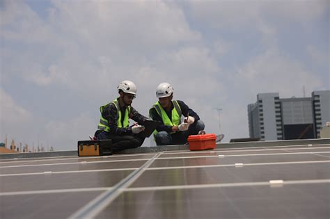 안전 조끼와 도구 상자를 착용한 전문 기술자가 햇빛 아래 공장 옥상에 태양광 지붕 패널을 설치하기 위해 확인합니다 엔지니어 팀