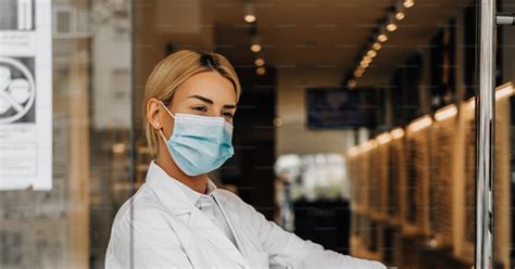 얼굴 보호 마스크를 쓴 젊고 매력적인 여성 검안사가 열린 안경점 문에 서서 밖을 내다보고 있습니다 그녀는 자신감 있고