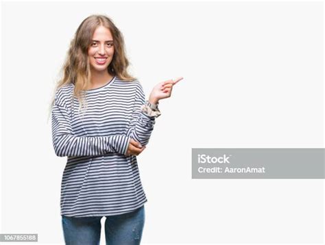 이상의 줄무늬 스웨터를 입고 아름 다운 젊은 금발 여자 카메라 보고 측면을 손과 손가락으로 가리키는 얼굴에 큰 미소와 함께 배경