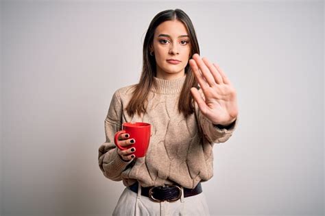 젊은 아름 다운 갈색 머리 여자 마시는 빨간 컵 의 커피 위에 고립 된 흰색 배경 와 열린 손 하 고 정지 기호 와 심각하 고 자신감 있는 표정 방어 제스처 금지됨에 대한