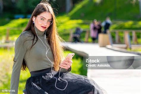 햇볕이 잘 드는 날에 휴대 전화를 사용하여 헤드폰으로 음악을 듣는 야외 공원에서 벤치에 앉아 있는 젊은 미소 짓는 여성 갈색 머리에 대한 스톡 사진 및 기타 이미지 Istock