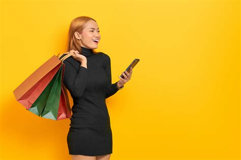 노란 배경을 통해 고립 된 복사 공간을 멀리 보고 쇼핑 백과 휴대 전화를 들고 미소 젊은 아시아 여성 25 29세에 대한 스톡 사진 및 기타 이미지 Istock