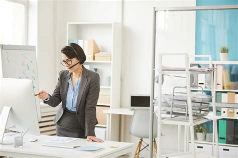 웹 회의 앱을 통해 원격 작업자를 직접 대면하여 연결하는 동안 테이블에 서서 모니터를 가리키는 핸즈프리 장치에서 자신감 있는 젊은 사업가 경영자에 대한 스톡 사진 및 기타