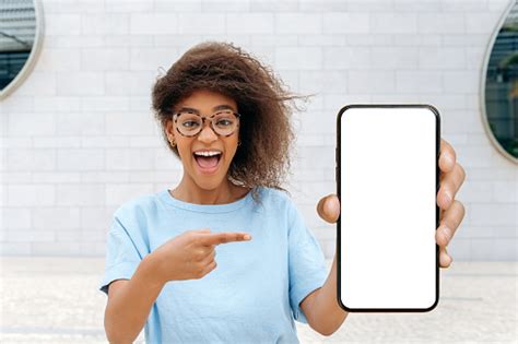 광고를위한 빈 공간 흥분한 충격에 놀란 아프리카 계 미국인 여성 야외에 서서 빈 흰색 모형 화면이있는 스마트 폰을 보여주고 손가락을 가리키고 카메라에 놀란 표정을 짓는다