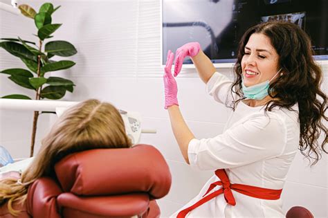 의자에 앉아 있는 환자에게 제스처를 가진 무언가를 보여주는 보호 장갑을 낀 채 턱에 의료용 마스크를 쓰고 있는 전문 치과 의사 여성 이빨에 대한 스톡 사진 및 기타 이미지