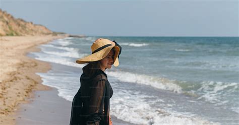 모자를 쓴 아름다운 평온한 여자는 바다 파도에 모래 해변을 걷고 휴식을 취합니다 여름 방학 밝은 검은 셔츠와 밀짚 모자를 쓴 세련된 젊은 여성이 열대 섬에서 휴가를