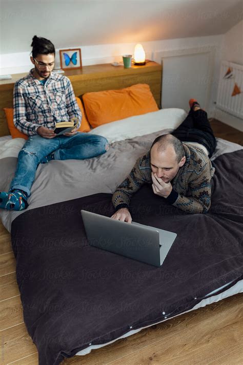 Young Gay Couple Relaxing On The Bed Reading A Book And Using A By