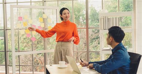 비즈니스 사람들은 사무실의 프로젝트 계획 보드에서 일하고 동료 친구와 대화하여 프로젝트 개발을 분석합니다 그들은 유리 벽에 게시 된 스티커 메모를 사용하여 정리합니다