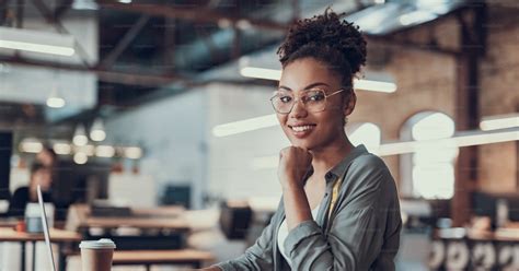 노트북과 문구류가 있는 나무 테이블에 앉아 있는 아름다운 젊은 아가씨의 초상화를 허리로 묶는다 그녀는 카메라를 바라보며 웃고 있는 동안 손에 턱을 괴고 있다 사진