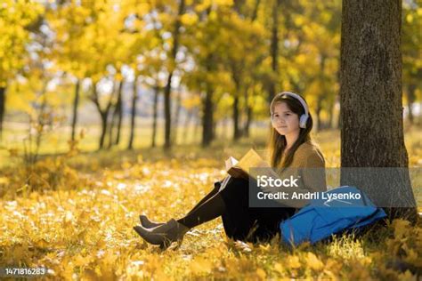 재미있는 책을 읽고 무선 헤드폰을 착용하고 화창한 가을날 공원의 나뭇잎에 앉아 있는 행복한 여학생 10 11세에 대한 스톡 사진 및 기타 이미지 Istock