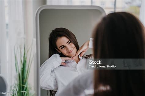 집에서 흰 셔츠를 입은 히스패닉의 멋진 젊은 여성은 거울을 바라보며 미소를 짓고 미용 절차 피부 관리 건강 및 미용 개념 후에 만족합니다 아름다움과 건강한 라이프 스타일