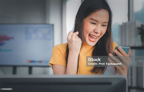 초상화 행복한 젊은 프로그래머 아시아 여성은 기술 창업 사무실에서 일하고 스마트 폰을 사용하고 모바일 응용 프로그램을 테스트하고 성공에 흥분합니다 똑똑한 기업가 여성의 개념