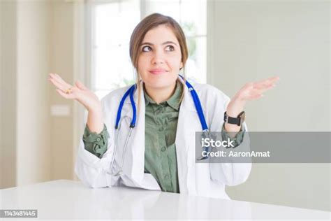 의료 코트와 청진 기를 쓰고 있는 아름 다운 젊은 의사 여자는 팔과 손을 제기 하 고 혼란 스러운 표정 의심의 개념입니다 감정에 대한 스톡 사진 및 기타 이미지 Istock