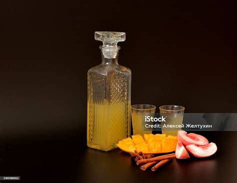 크리스탈 디캔터에 담긴 복숭아 망고 리큐어와 검은 배경에 두 잔의 잔 그 옆에는 계피 스틱 아니스 잘 익은 과일 조각이 있습니다 검정색 배경에 대한 스톡 사진 및 기타