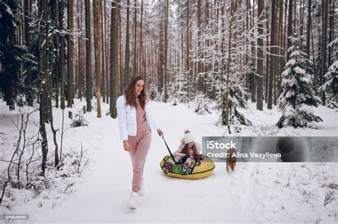 행복한 어머니와 핑크 따뜻한 아웃웨어를 입은 어린 귀여운 소녀는 야외에서 눈 덮인 흰색 겨울 숲에서 붉은 시바 이누 개와 함께 풍선 스노우 튜브를 재미있게 타는 것을