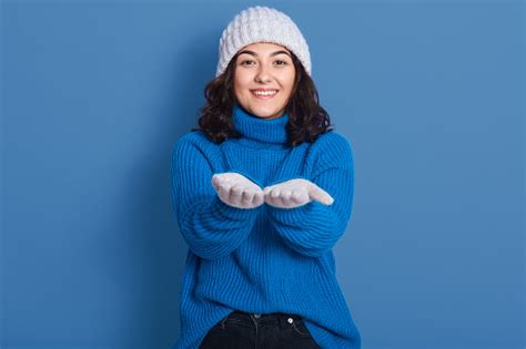 파란색 스튜디오 배경에 고립 된 포즈를 취한 사랑스러운 여성의 클로즈업 초상화 흰색 모자와 스웨터를 입고 두 손바닥을 함께 유지하고 카메라에 보여주는 것은 행복하고 기쁘게