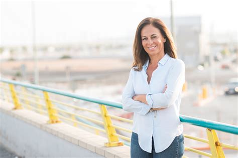 아름 다운 중 년 스페인 여자는 거리에서 얼굴에 미소와 함께 서 있는 자신감과 밝은 화창한 날에 웃 고 있는 동안 도시를 걷고 갈색 머리에 대한 스톡 사진 및 기타 이미지