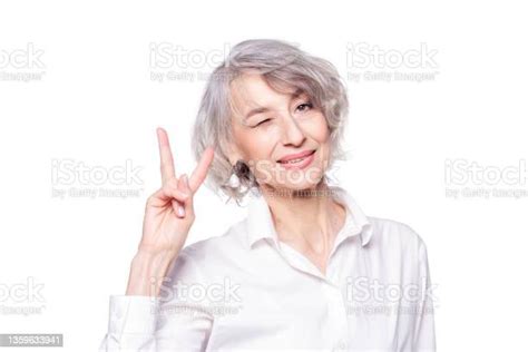 편안한 흰색 배경 위에 서있는 우아한 셔츠를 입은 선임 회색 머리 여성의 초상화를 클로즈업하여 승리의 사인을하는 카메라에서