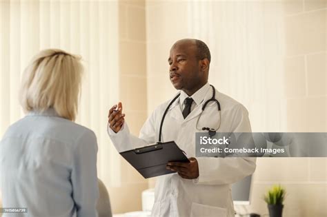 여성 환자에게 진단을 설명하는 클립 보드가있는 흰색 코트의 흑인 의사 의사 사무실에서 일반 개업의의 말을 듣는 인식 할 수없는 금발 여성 25 29세에 대한 스톡 사진 및