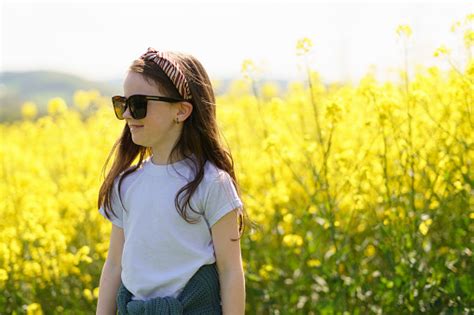 흰 티셔츠에 큰 선글라스를 끼고 허리띠에 스웨터를 묶은 쾌활한 소녀가 노랗게 피어난 유채밭 한가운데에 쾌활하게 서 있습니다 아이들의 감성과 여가의 개념 6 7 살에 대한