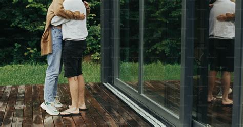 세련된 힙스터 커플은 나무 테라스에서 포옹하고 키스하며 산의 오두막에서 휴식을 취합니다 큰 창문과 나무를 배경으로 현대적인 의상을 입은 행복한 젊은 가족 사진