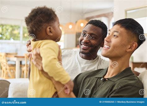 Same Sex Male Couple Or Friends Playing With Baby Sitting On Sofa At Home Together Stock Image
