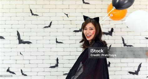 할로윈 파티에서 검은 종이 박쥐 모양으로 장식 된 흰색 벽돌 배경에 풍선으로 포즈를 취하는 마녀 의상을 입은 젊은 아름다운 아시아 여성의 초상화 복사 텍스트를위한 여유 공간