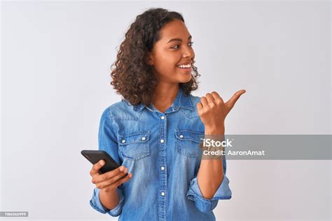고립 된 흰색 배경 위에 서서 행복한 얼굴미소를 지으며 엄지 손가락으로 보여주는 스마트 폰을 사용하는 젊은 브라질 여성 가리키기에 대한 스톡 사진 및 기타 이미지 Istock