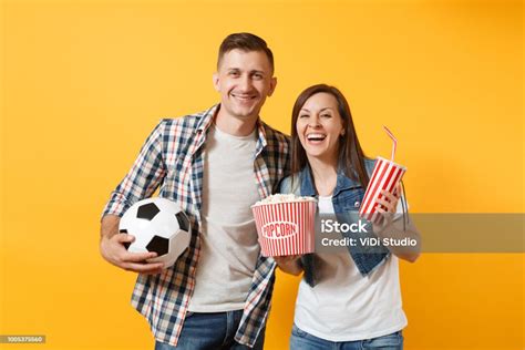 젊은 커플 여자 남자 축구 팬 들 들고 축구공 팝콘의 물통 플라스틱 유리 콜라의 재미 지원 팀 노란 배경에 고립 된 기운 스포츠 가족 레저 라이프 스타일 개념입니다