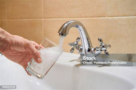 스테인레스 스틸 기둥 탭에서 물 한 잔을 채우는 남자 화장실 크롬 수도꼭지에서 물을 유리에 붓는 남성의 손이 기포로 흐르는 물을 마십니다 식수 와 안전 음료 가정 생활에
