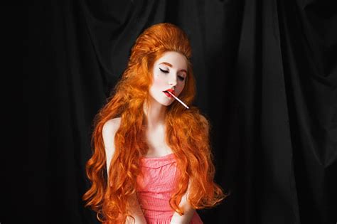 창백한 피부와 검은색 바탕에 핑크 드레스에 긴 곱슬 붉은 머리 흡연 여자 그녀의 손에 담배와 붉은 입술으로 아름 다운 여자 뷰티 메이크업입니다 빨간 머리 모델입니다 관능적인