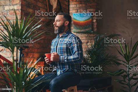 블루 양 털 셔츠와 청바지에 잘생긴 수염된 Hipster 남성 다락방 인테리어 스튜디오에서 나무의 자에 앉아있는 동안 아침 커피 한 잔을 보유 하고있다 가을에 대한 스톡
