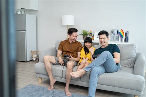 아시아 게이 커플 놀이 비디오 게임 와 어린 딸 에 거실 작은 사랑스러운 소녀 아이 앉아 소파에 매력적인 로맨틱 한 젊은 남성 Lgbtq 가족 조이스틱을 사용 하 여 즐길