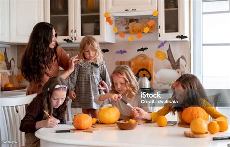 귀여운 젊은 어머니는 행복한 어린 딸들과 함께 집에있는 밝은 흰색 부엌에서 호박에 유령 같은 얼굴을 조각하는 칼을 들고 있습니다 할로윈 파티 준비 부엌에 대한 스톡 사진 및