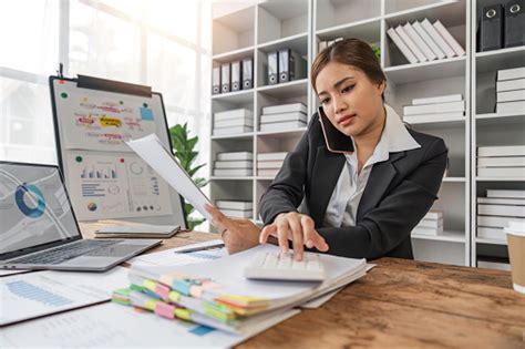 비즈니스 우먼은 노트북 앞 사무실 책상에 앉아 휴대폰을 들고 즐거운 비즈니스 또는 비공식 통화를 한다 계산기로 재정을 계산하는 성공적인 사업가 경영자에 대한 스톡 사진 및