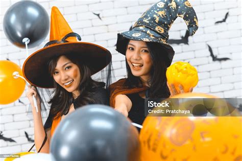 할로윈 파티를위한 검은 종이 박쥐 모양으로 장식 된 흰색 벽돌 배경에 풍선을 들고 마녀 할로윈 의상을 입은 두 명의 아름다운 젊은 아시아 여성 공휴일에 대한 스톡 사진 및