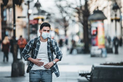 젊은 남자는 마을에 서있는 동안 얼굴 마스크와 기침을 착용합니다 의료 얼굴 마스크와 젊은 남자의 초상화는 도시 거리에 서있다 사람들이 군중에서 야외 바이러스에서 보호