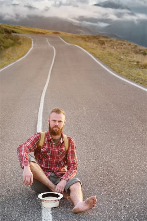 Stylish Barefoot Bearded Male Hitchhiker Traveler In A Hat And With A Backpack Sits On A