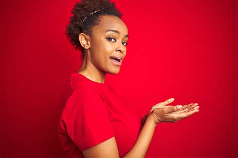 고립 된 빨간 배경 위에 아프리카 머리를 가진 젊은 아름다운 아프리카 계 미국인 여성은 복사 공간을 보여주는 손바닥을 옆으로 가리키며 행복한 미소를 짓는 광고를 제시합니다