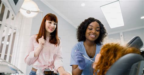 검진 중 치과 진료소에서 어린 딸을 지원하는 젊은 여성 엄마 파란색 유니폼을 입고 장갑을 낀 아프리카 소아 치과 의사가 어린 소녀의 치아를 검사하고 있다 사진