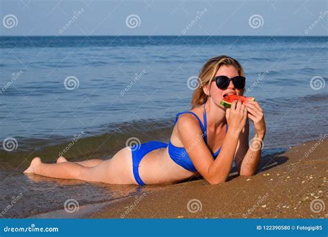 The Blonde In Sunglasses Lies On The Beach Near The Waves And Eats A